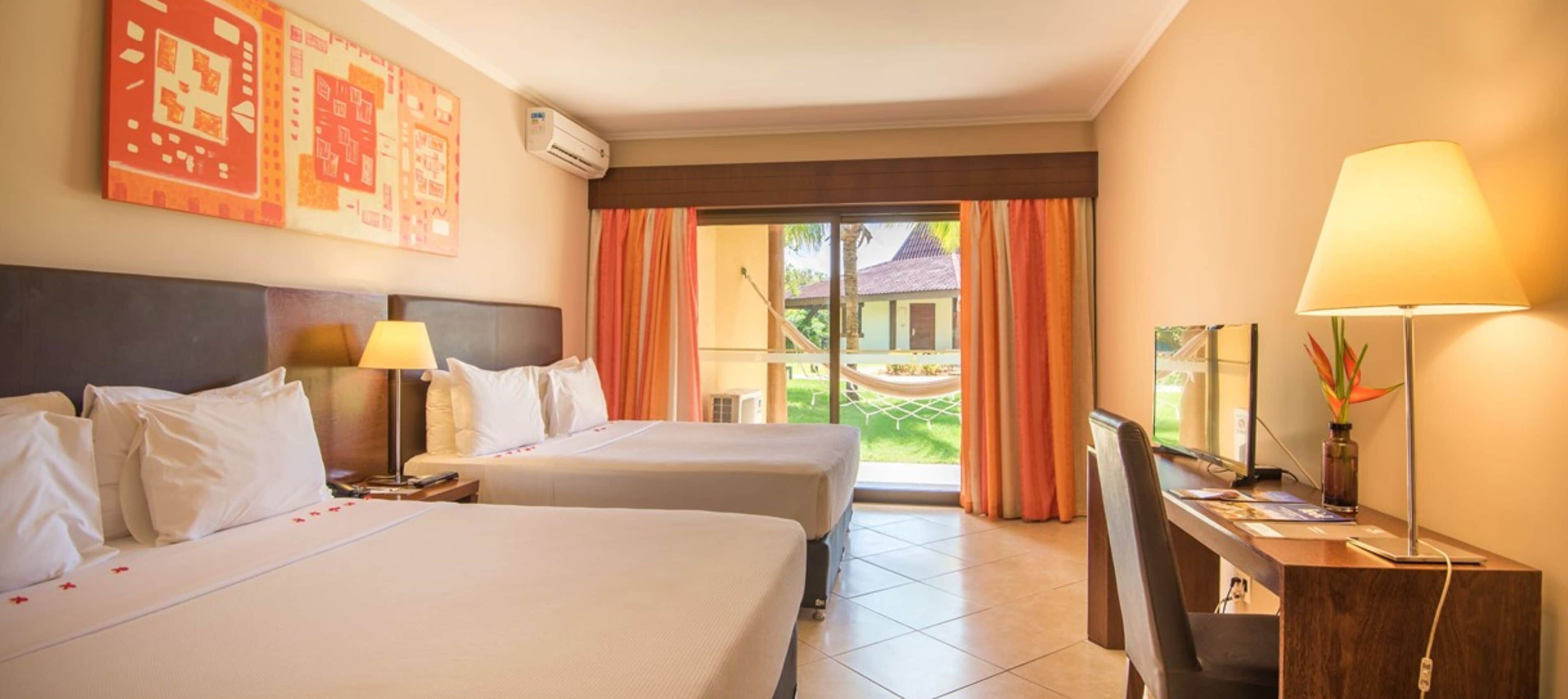 Quarto de hotel com duas camas, uma escrivaninha, uma cadeira, uma luminária e vista para o exterior.