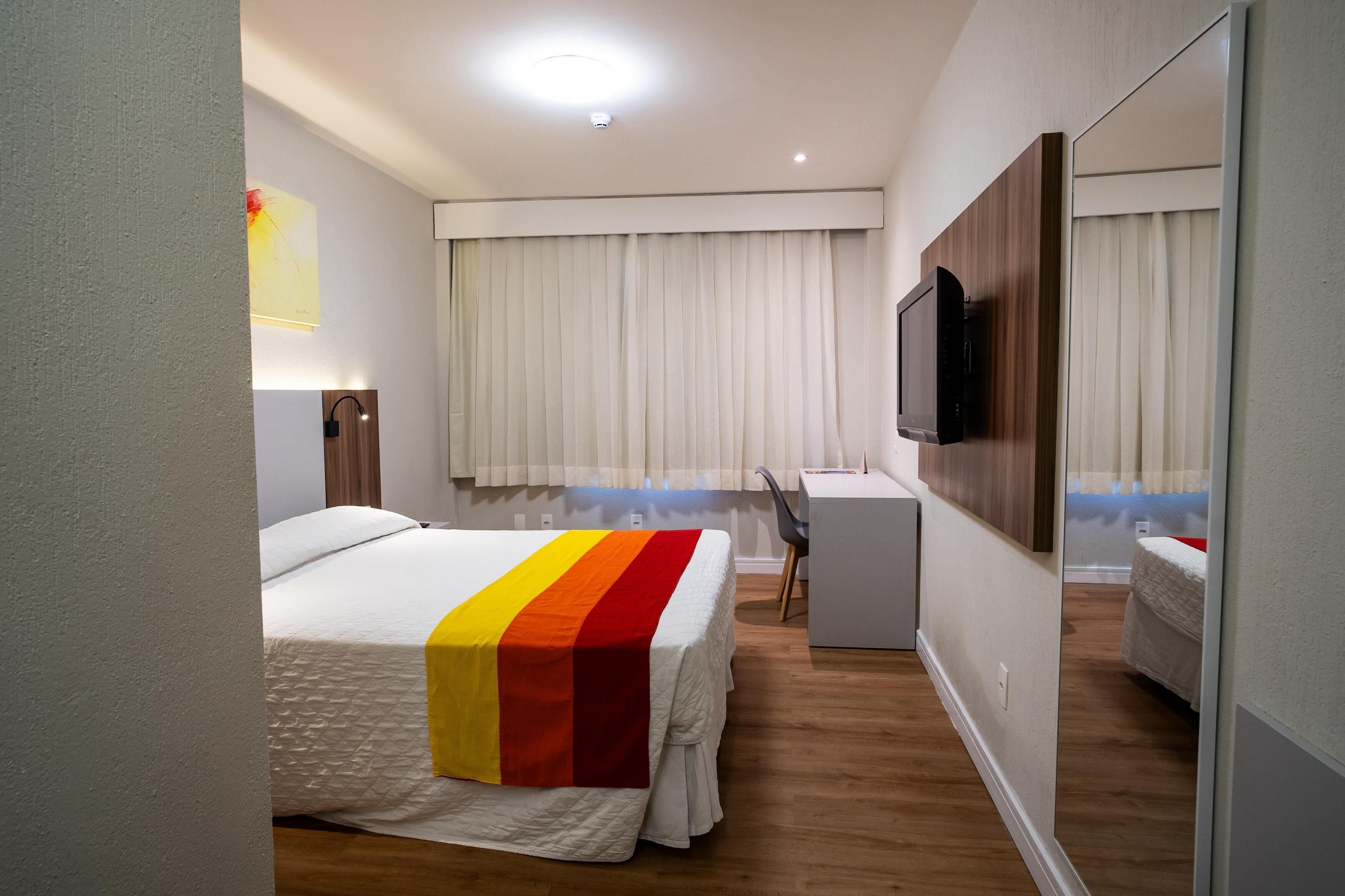Quarto de hotel com cama, escrivaninha, cadeira, televisão e janela com cortinas.