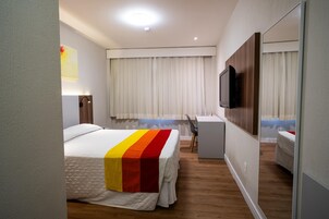 Quarto de hotel com cama, escrivaninha, cadeira, televisão e janela com cortinas.