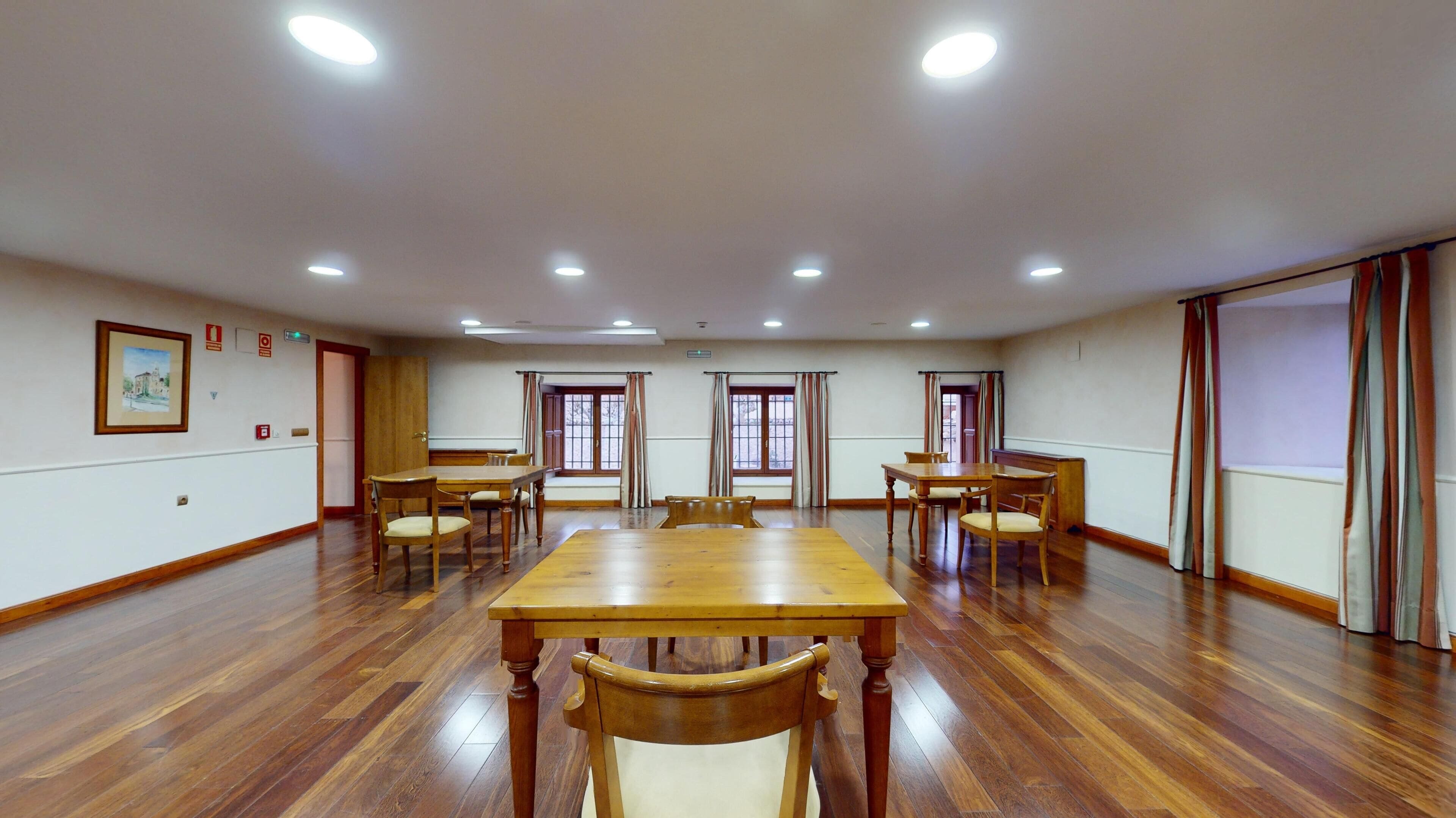 Um quarto espaçoso com uma grande mesa de madeira e cadeiras, iluminação embutida no teto, janelas com cortinas e um quadro na parede.