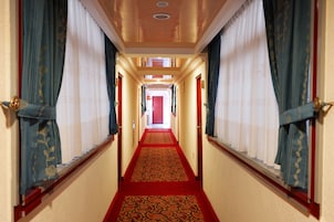 Um corredor de hotel com carpete vermelho, cortinas azuis estampadas com flores e uma porta vermelha no final.