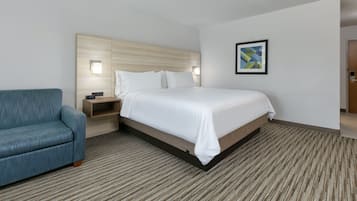 Quarto de hotel com uma cama, uma poltrona azul, uma mesa de cabeceira e um quadro na parede.
