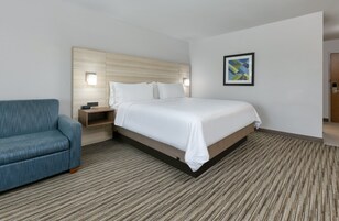 Quarto de hotel com uma cama, uma poltrona azul, uma mesa de cabeceira e um quadro na parede.