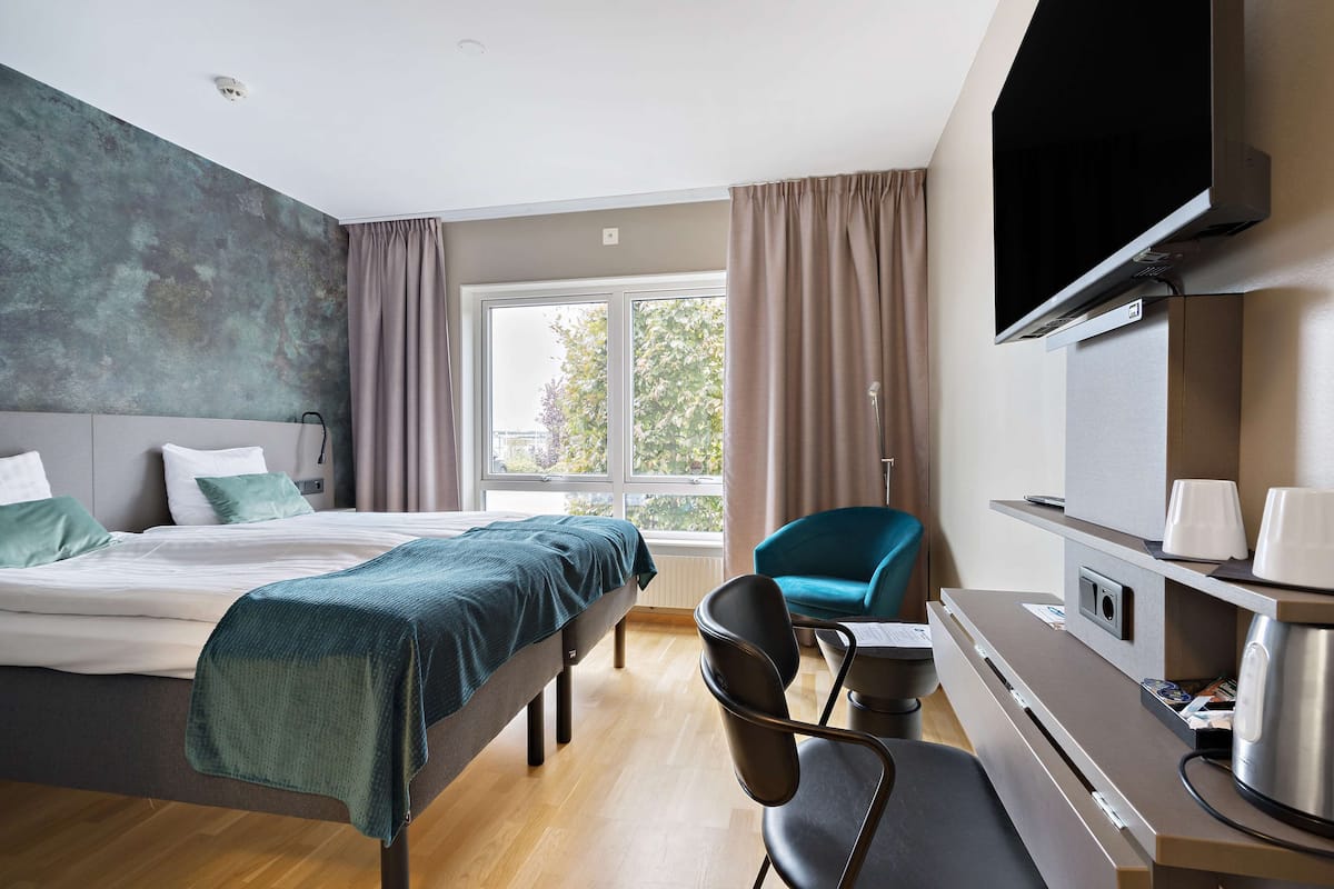 Quarto de hotel moderno com cama, escrivaninha, cadeira, televisão e janela com cortinas.