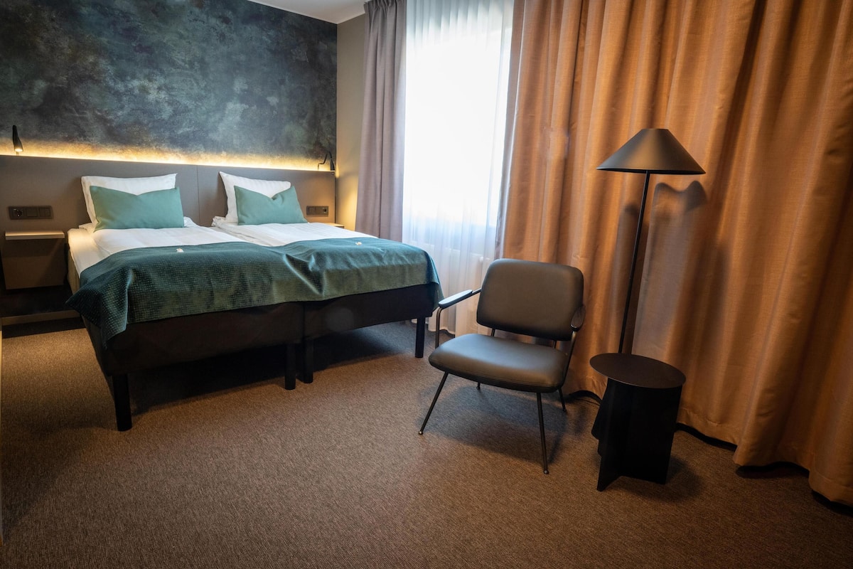 Quarto de hotel com cama, cadeira, mesa pequena, abajur e uma janela grande com cortinas.