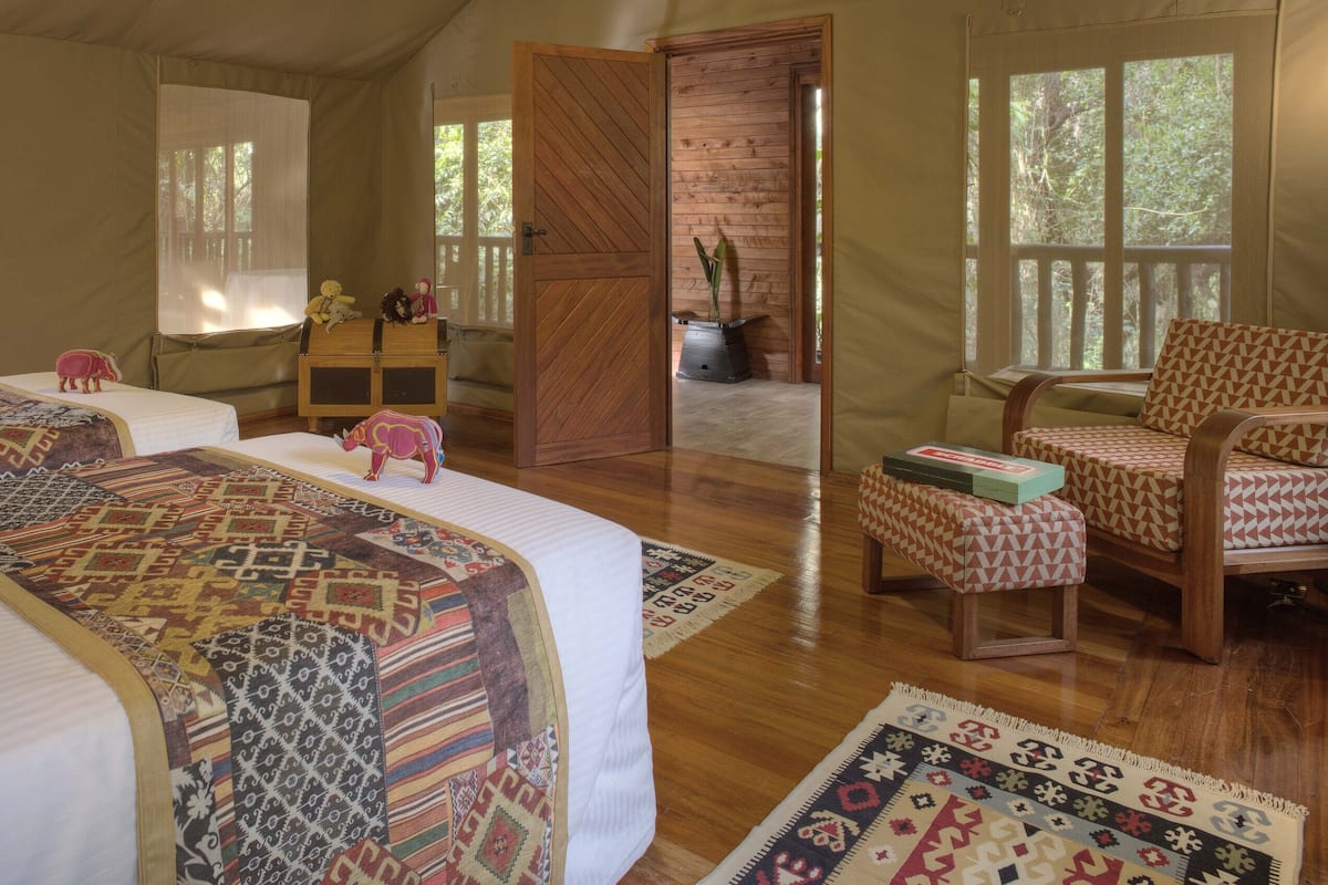 Quarto acolhedor com duas camas, uma cadeira de madeira, uma mesa pequena e janela com vista para árvores.