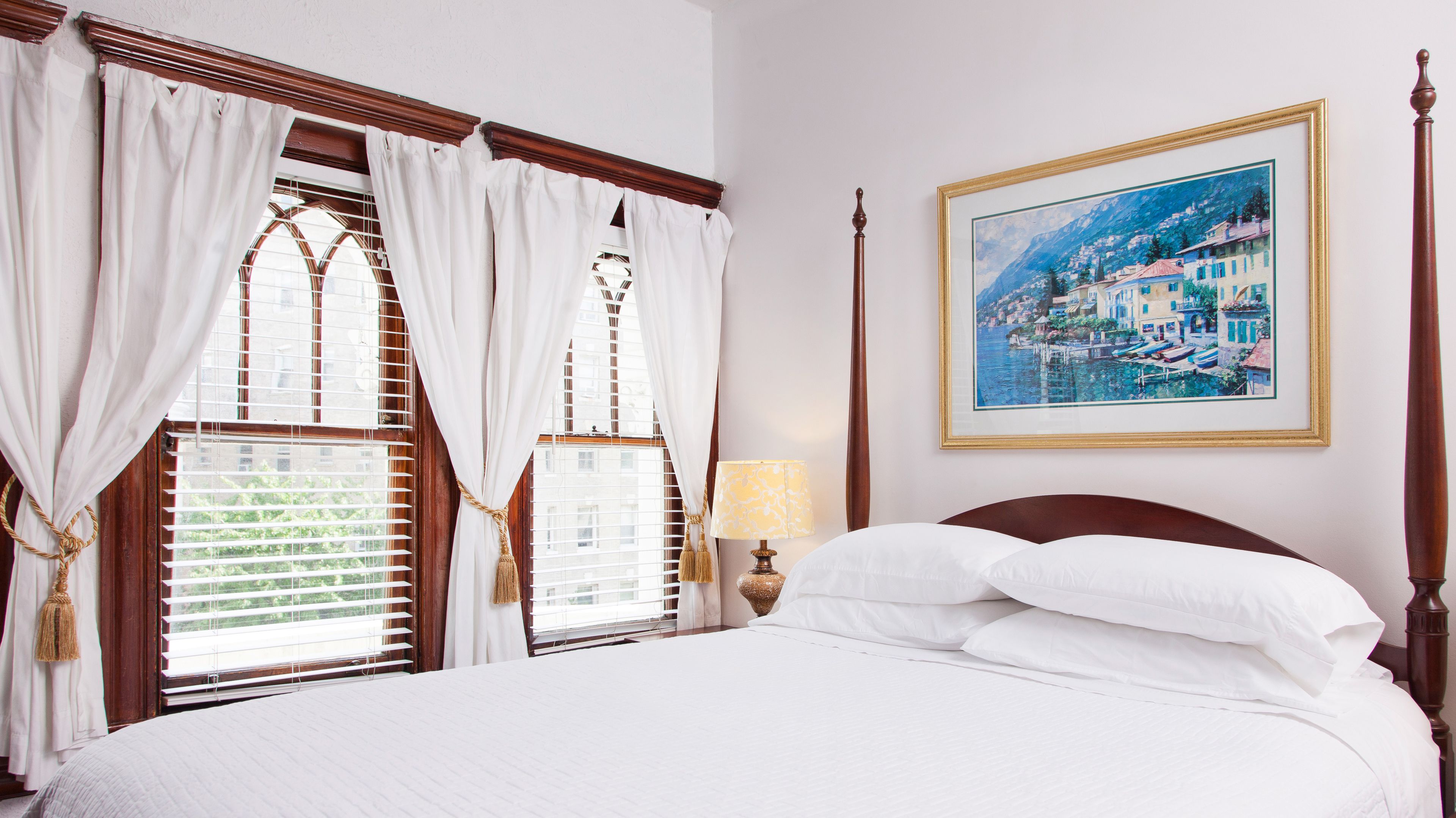 Cama bem arrumada com lençóis brancos, cabeceira de madeira e um quadro de uma cidade costeira na parede.