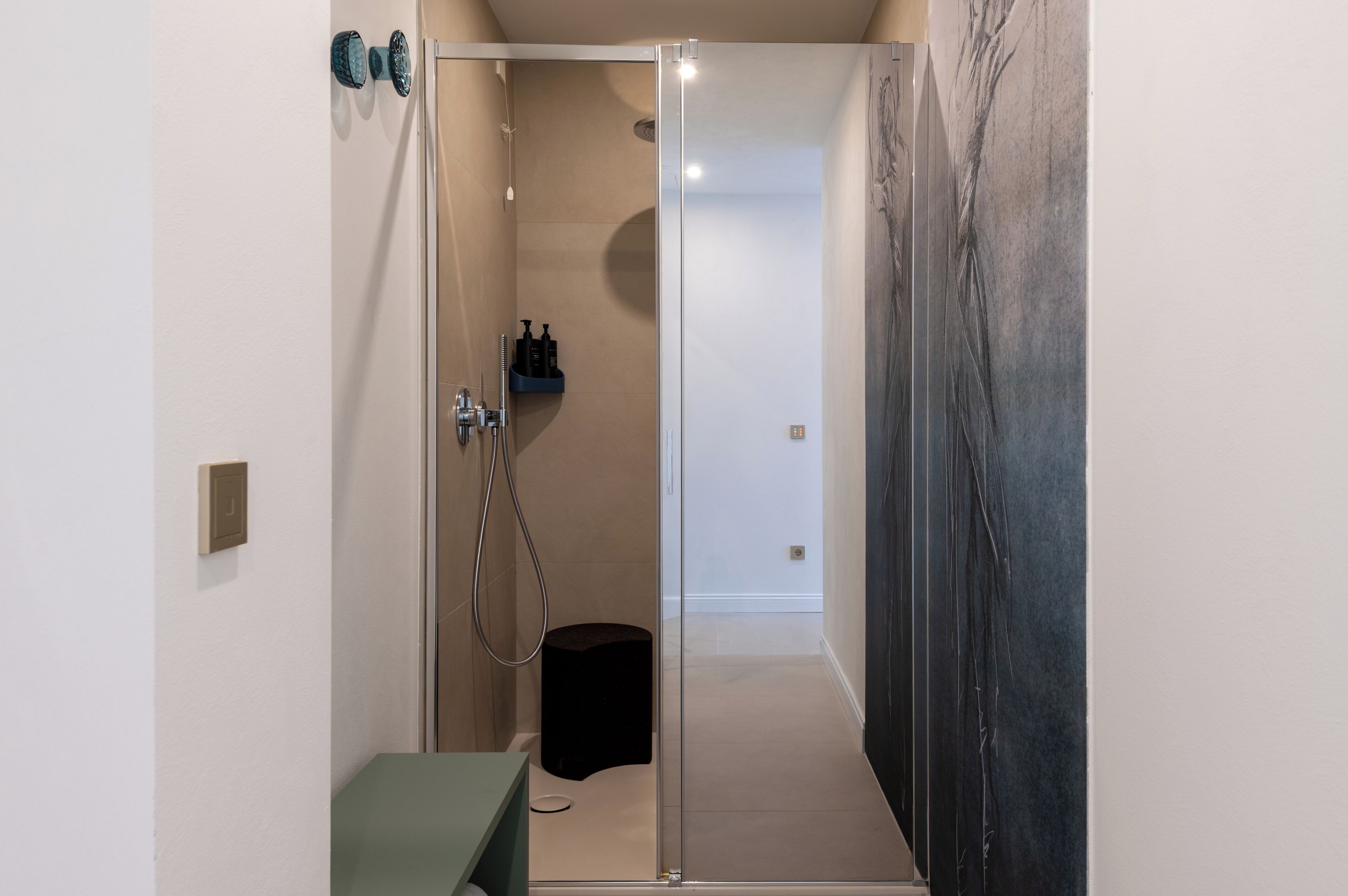 Casa de banho moderna com cabine de chuveiro em vidro, um banquinho pequeno e uma prateleira com produtos de higiene pessoal.