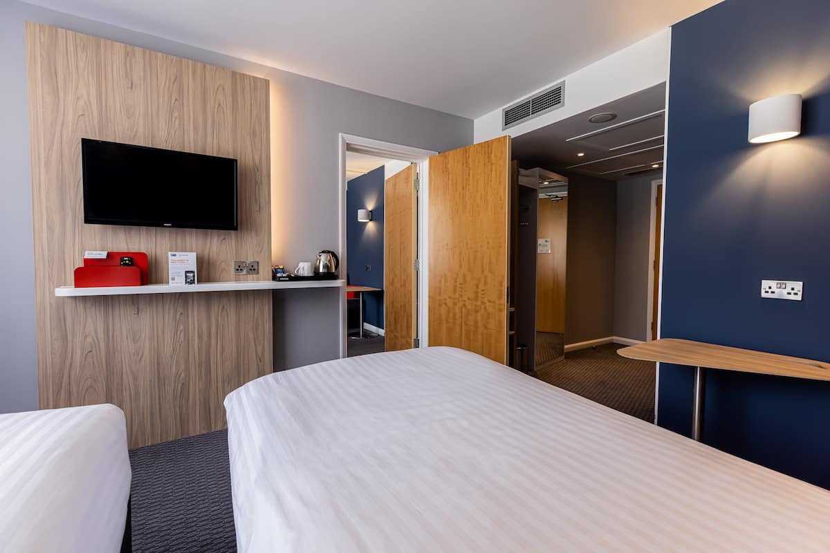 Quarto de hotel com cama, televisão fixada na parede, pequena mesa com cadeira vermelha e painel de parede em madeira.