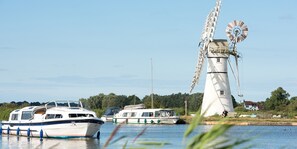 Um moinho de vento tradicional à beira de um rio calmo, com barcos atracados por perto.
