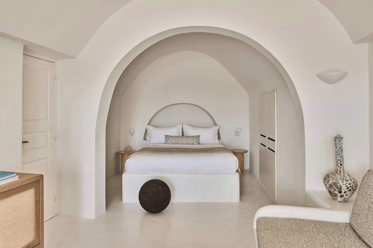 Quarto com uma grande porta em arco, cama com roupa de cama branca, um banquinho redondo, uma cadeira branca e um vaso decorativo.