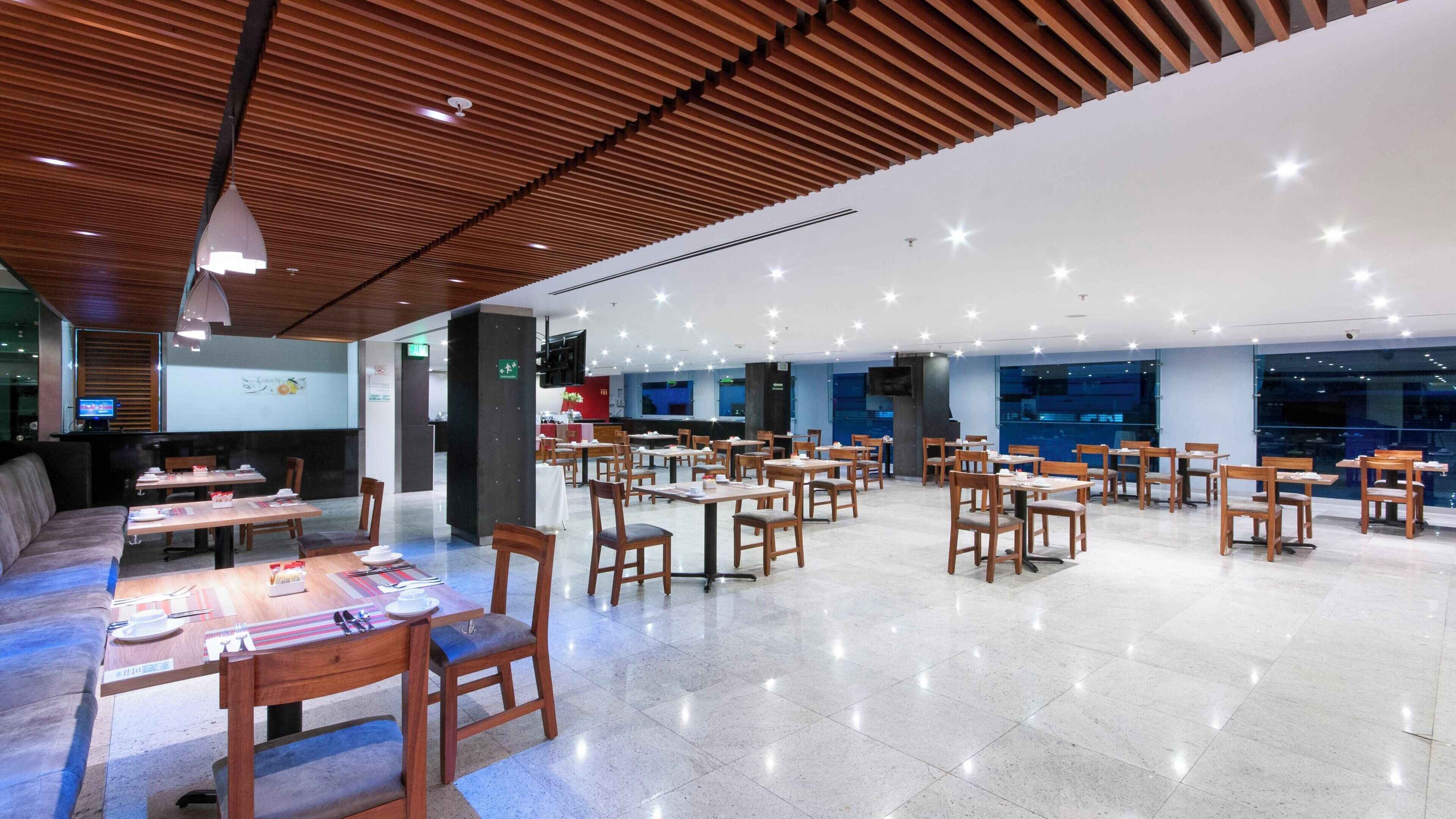 Um restaurante amplo com pé-direito alto, vigas de madeira e design moderno. Mesas e cadeiras estão organizadas, e o chão é revestido com azulejos.
