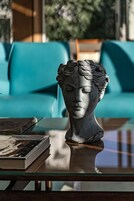 Escultura de busto sobre uma mesa de vidro com um livro ao lado, em um quarto com sofás verde-água e uma janela.