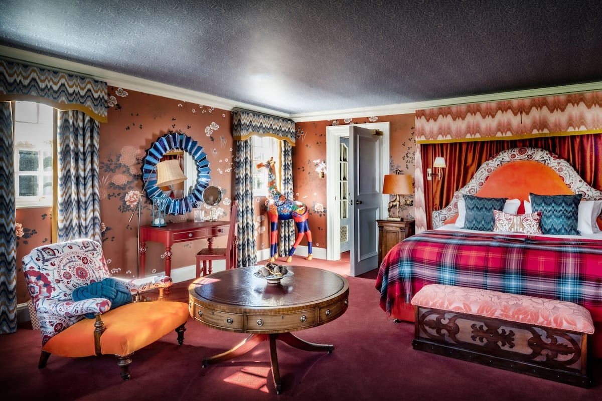 Quarto com uma mesa redonda de madeira, uma cama com colcha xadrez e uma cadeira com almofada estampada.