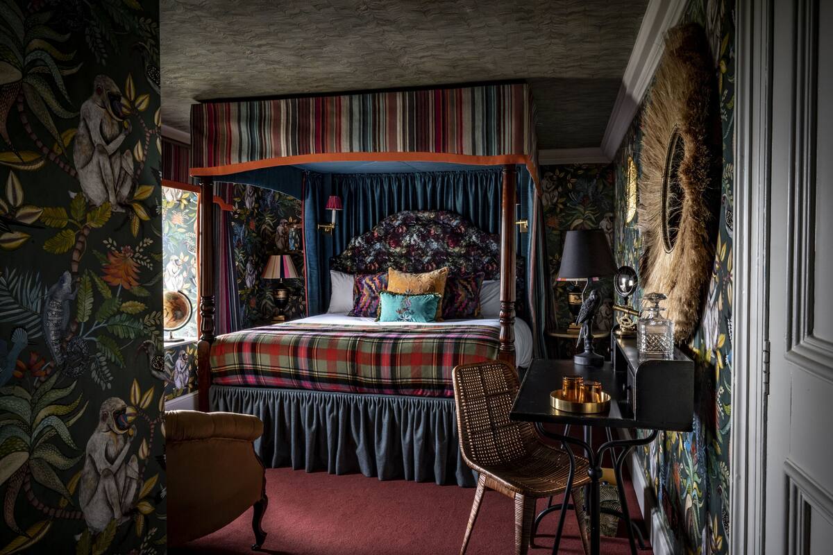 Uma cama com dossel com colcha xadrez, uma cadeira de madeira e uma pequena mesa com abajur e objetos decorativos.