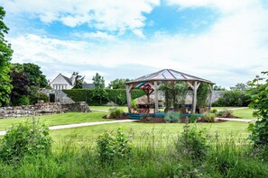 Um gazebo com base de pedra e teto de metal, rodeado por um jardim bem cuidado com gramado e diversas plantas.