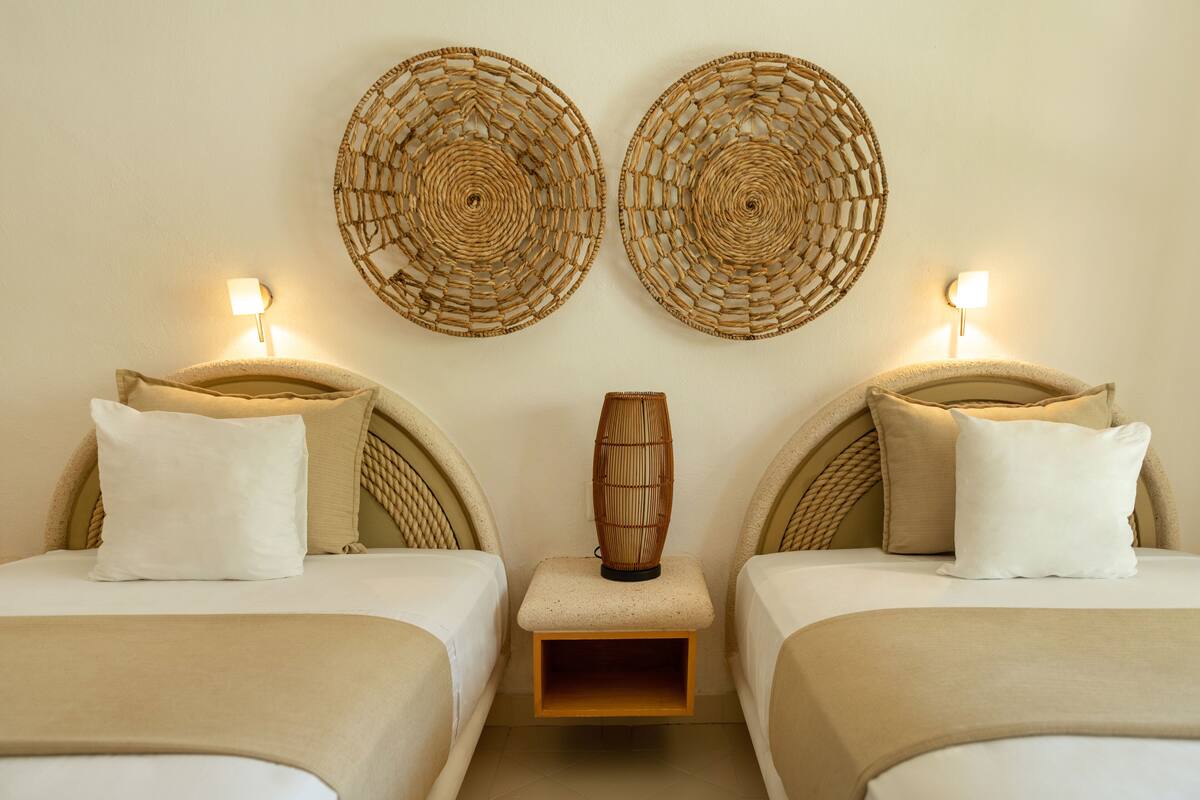 Quarto de hotel com duas camas, cada uma com almofada e uma pequena mesa de cabeceira com abajur.