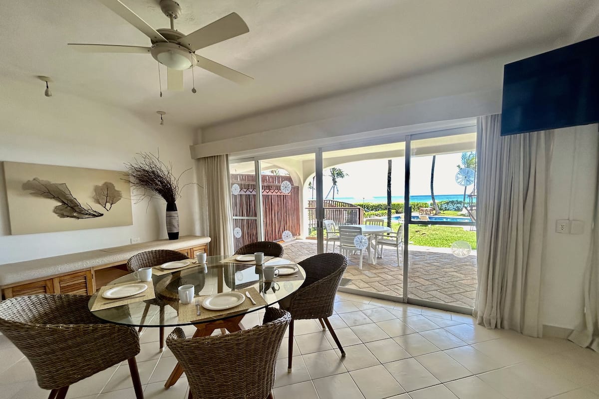 Uma sala de jantar com mesa de vidro, cadeiras de vime e vista para uma piscina e praia.