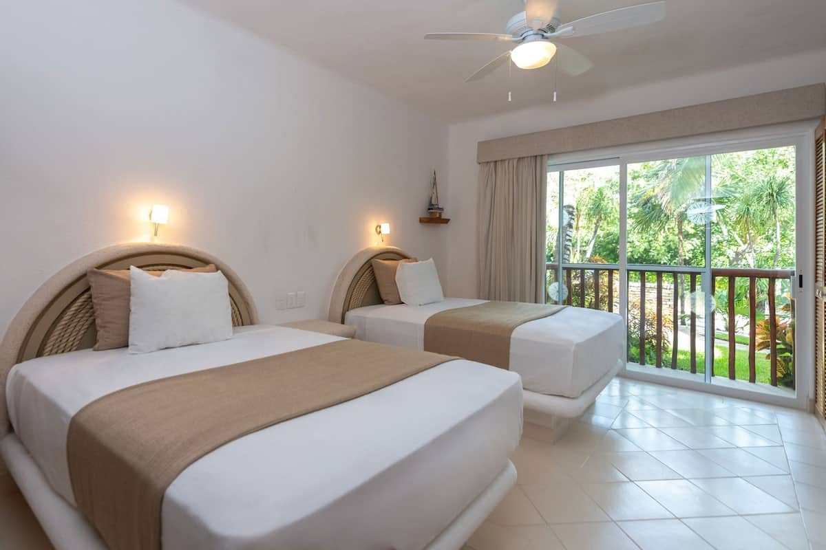 Quarto de hotel com duas camas, um ventilador de teto, uma janela grande com vista para áreas verdes e uma varanda.