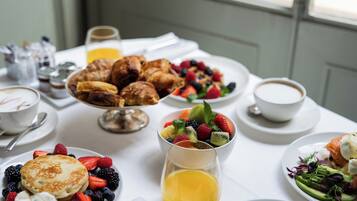 Uma mesa de pequeno-almoço com vários pratos, incluindo panquecas, croissants e uma salada de frutas, posta sobre uma toalha de mesa branca.