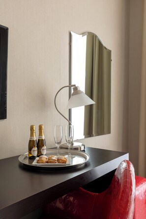 Quarto de hotel com escrivaninha de madeira escura, uma luminária, duas garrafas de champanhe, dois copos e um prato com doces.