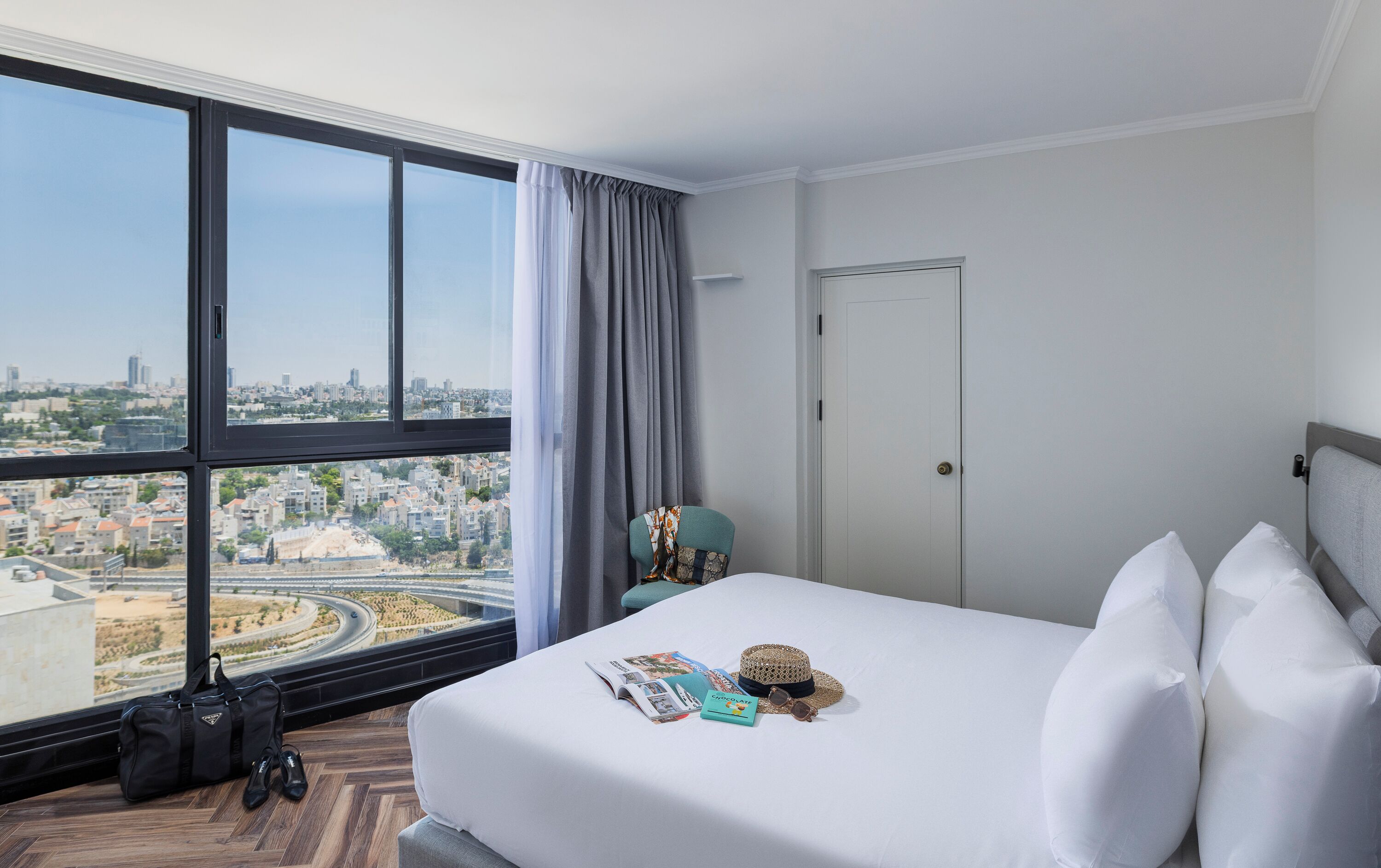 Quarto de hotel com uma grande janela que oferece vista para a cidade, uma cama bem arrumada, uma cadeira e uma mala no chão.