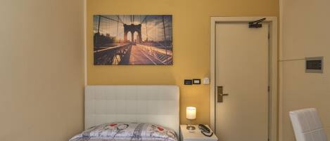 Quarto de hotel com cama, mesa de cabeceira, abajur, porta e um quadro na parede.