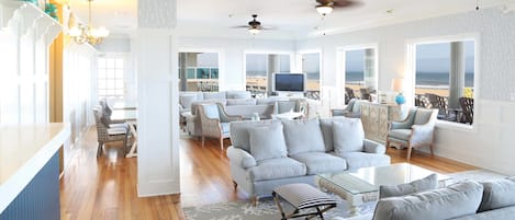 Uma sala de estar espaçosa com um sofá grande, uma mesa de centro e vista para a praia. Há ventiladores de teto e uma TV no ambiente.