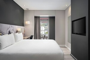 Quarto de hotel moderno com uma cama grande, uma televisão de tela plana, uma mesinha com cadeiras e vista para a cidade através de uma porta de vidro.