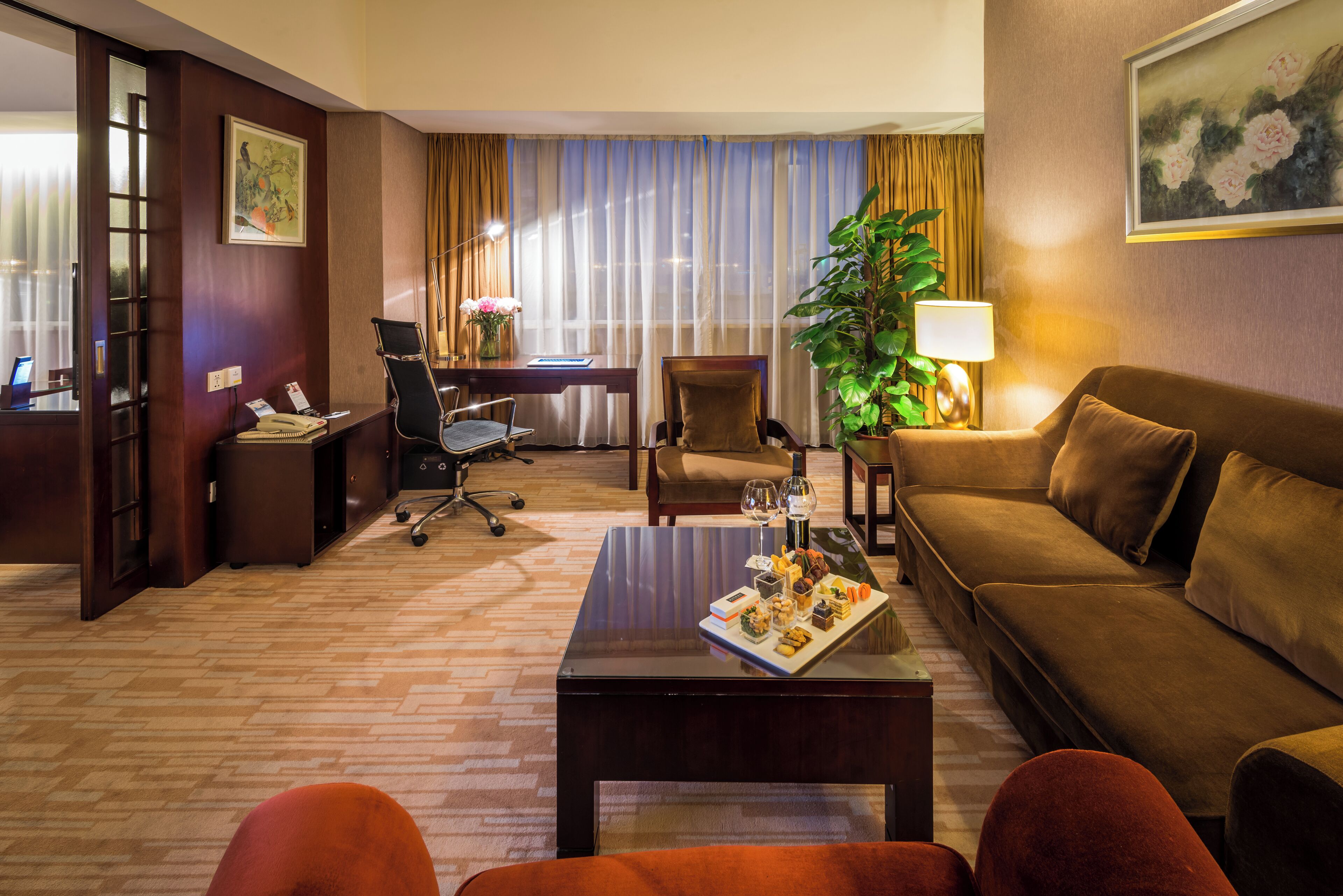 Quarto de hotel com piso de madeira, sofá, mesa de centro com petiscos, escrivaninha com cadeira e uma grande janela com cortinas.