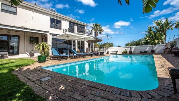 Piscina com espreguiçadeiras e guarda-sóis, casa com varanda e plantas tropicais.