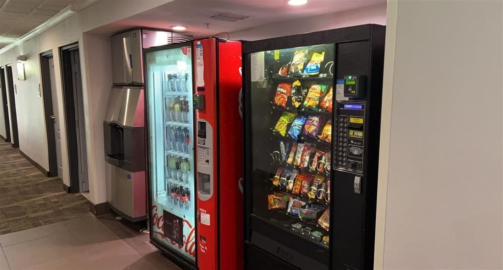 Corredor com máquinas de venda automática, incluindo um refrigerador de Coca-Cola e um distribuidor de lanches.