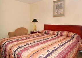 Quarto de hotel com uma cama, uma cadeira, uma mesinha de cabeceira, um abajur e um quadro pendurado na parede.