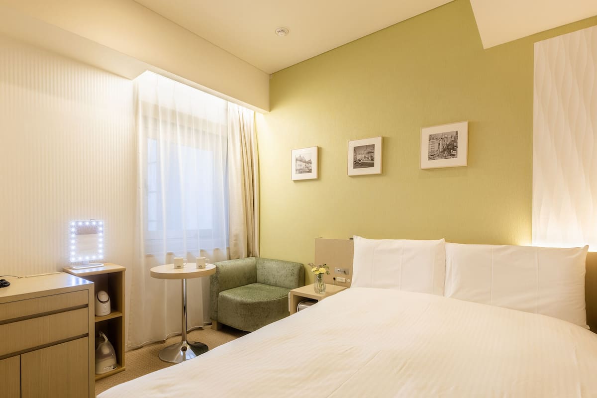 Quarto de hotel com uma cama, uma penteadeira com espelho, uma mesa redonda pequena com duas xícaras, uma poltrona verde e três quadros na parede.