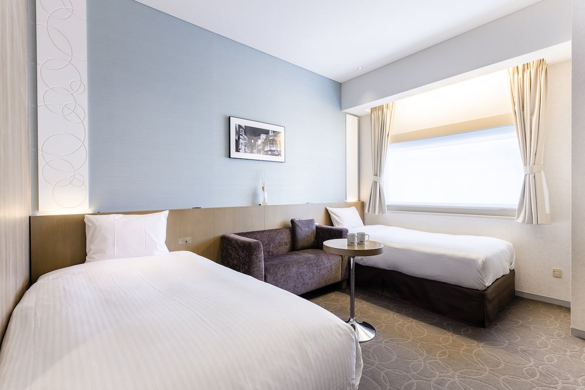 Quarto de hotel com duas camas, um sofá, uma mesa pequena e uma janela com cortinas.