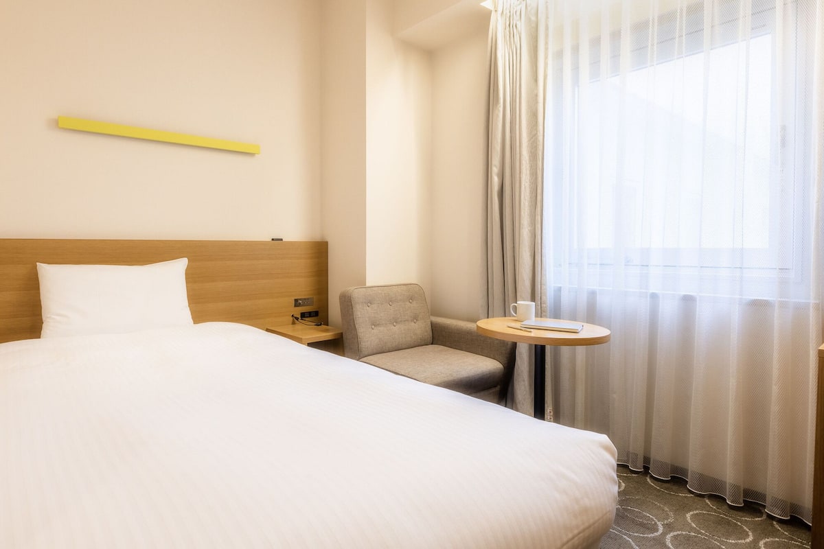 Quarto de hotel com uma cama grande, uma poltrona pequena, mesa de cabeceira com abajur e vista para o exterior através de cortinas translúcidas.