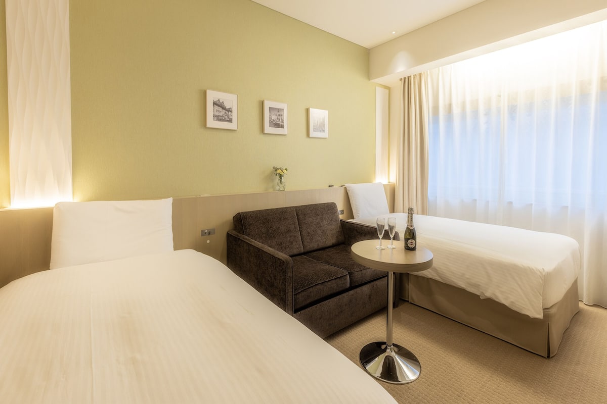 Quarto de hotel com duas camas, um sofá pequeno, uma mesa redonda com uma garrafa e copos, e uma janela com cortinas.