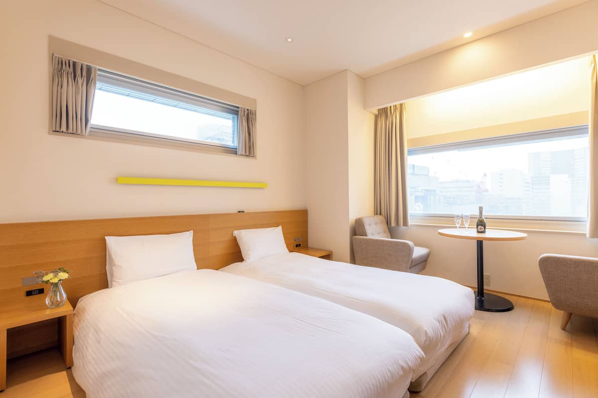 Quarto de hotel com duas camas, uma mesa pequena com dois copos, uma cadeira e uma janela com cortinas.