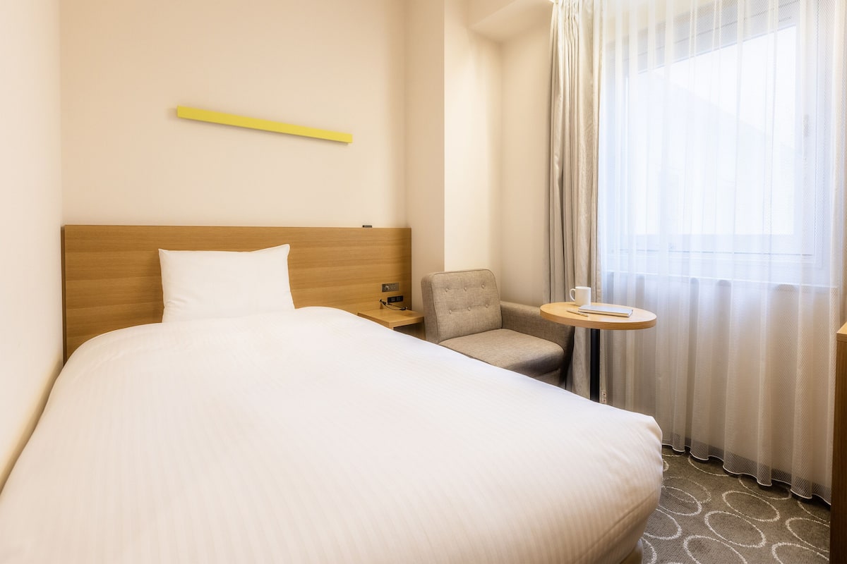 Quarto de hotel com uma cama grande, uma poltrona pequena, uma mesinha de cabeceira e uma janela com cortinas finas.