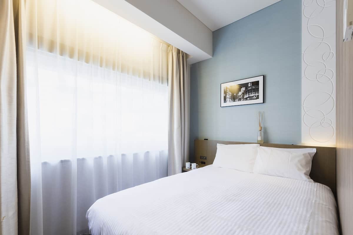 Quarto de hotel com uma cama grande, uma mesa de cabeceira com abajur, um quadro na parede e cortinas translúcidas na janela.