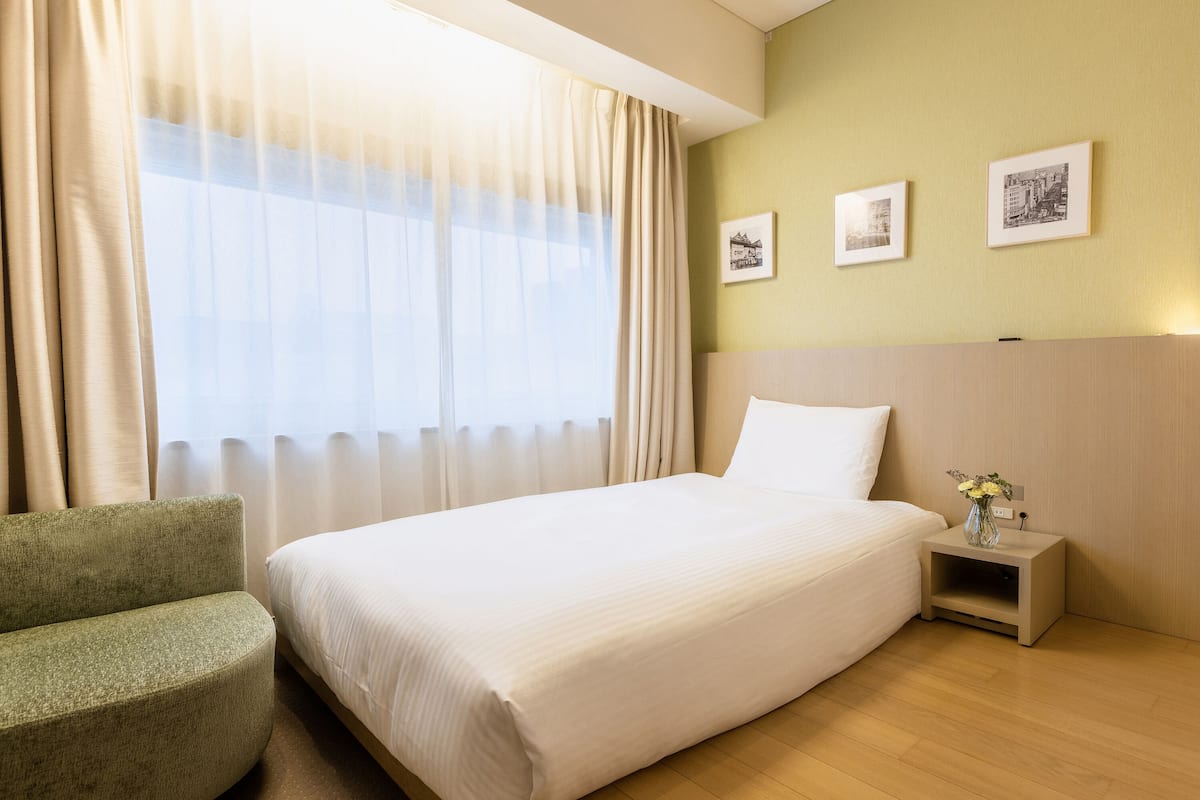 Quarto de hotel com uma cama grande, uma poltrona verde, uma mesa de cabeceira com um vaso de flores e três quadros na parede.
