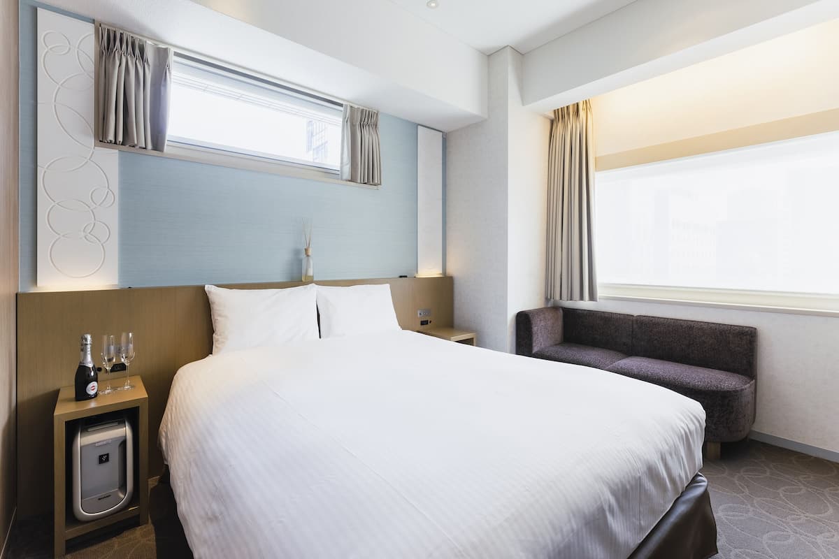 Quarto de hotel moderno com uma cama grande, mesa de cabeceira com minibar, área de estar e uma janela ampla com cortinas.