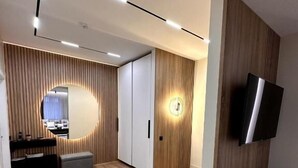 Quarto de hotel moderno com piso de madeira, um espelho redondo, uma TV de tela plana na parede e um pequeno banco.