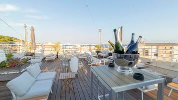Terraço no telhado com cadeiras de lounge brancas, uma mesa com garrafas e vista ampla da cidade.