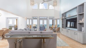Ampla sala de estar com um sofá cinza grande, uma mesa de centro de vidro e uma TV montada em um armário de madeira. Há várias janelas com vista para o oceano, e o cômodo possui um pé-direito alto com ventilador de teto.