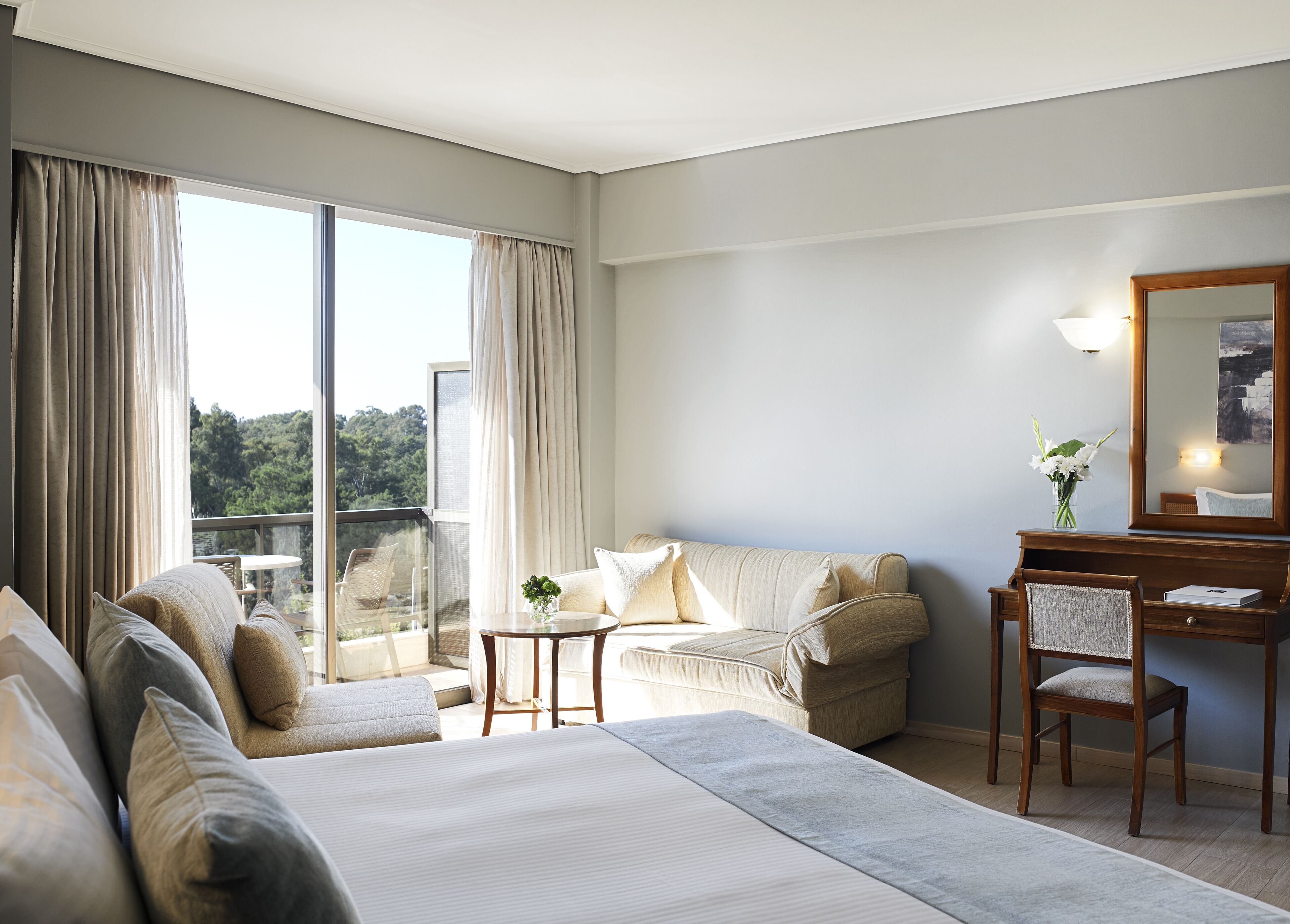 Quarto de hotel com cama, escrivaninha, cadeira e uma grande janela com vista para o verde.
