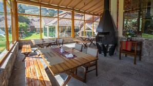 Um alpendre exterior com mesas e cadeiras de madeira, uma parede de pedra e vista para uma aldeia com casas e vegetação.