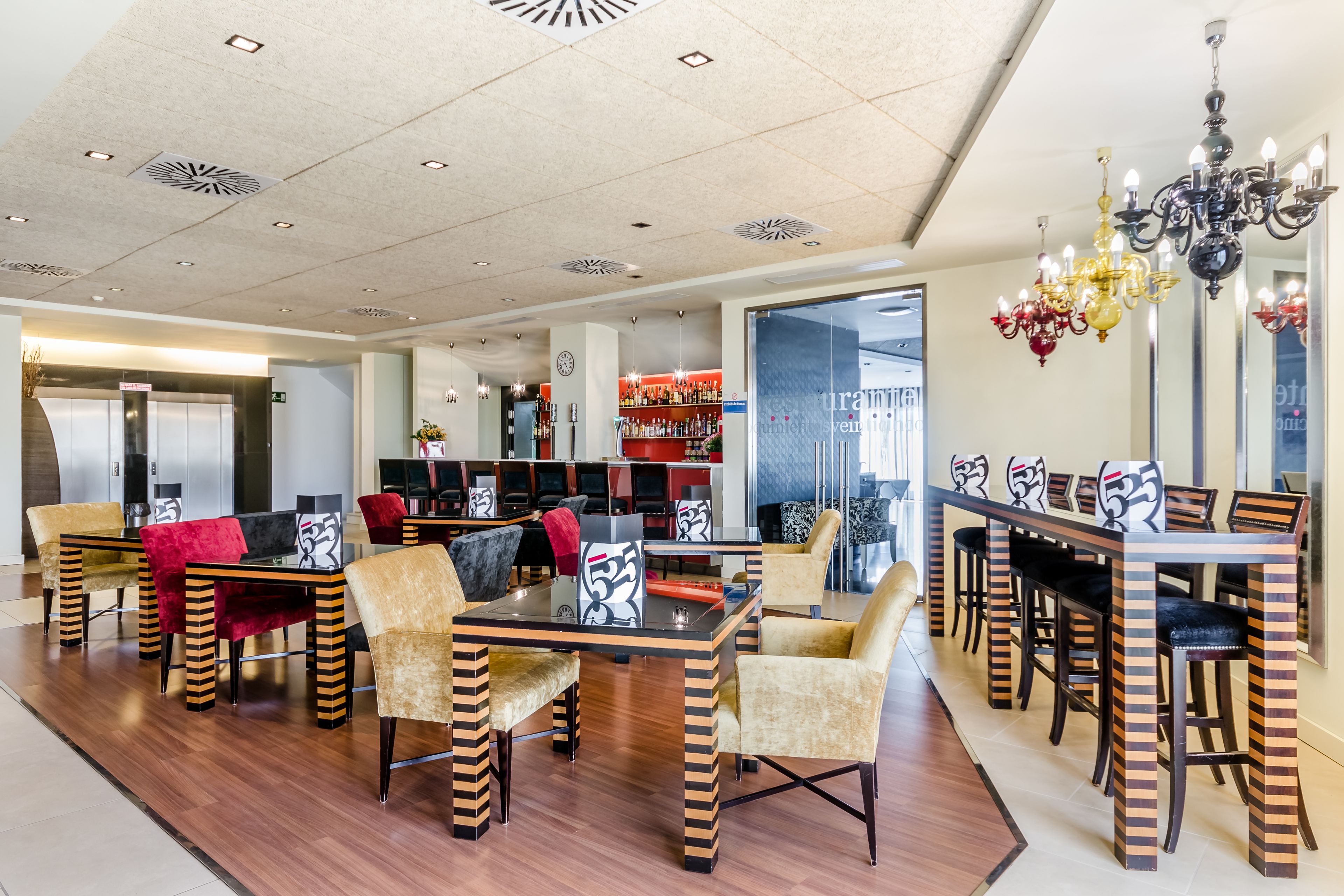 Uma ampla área de jantar com cadeiras e mesas listradas, um espaço de bar com prateleiras cheias de garrafas e um lustre.