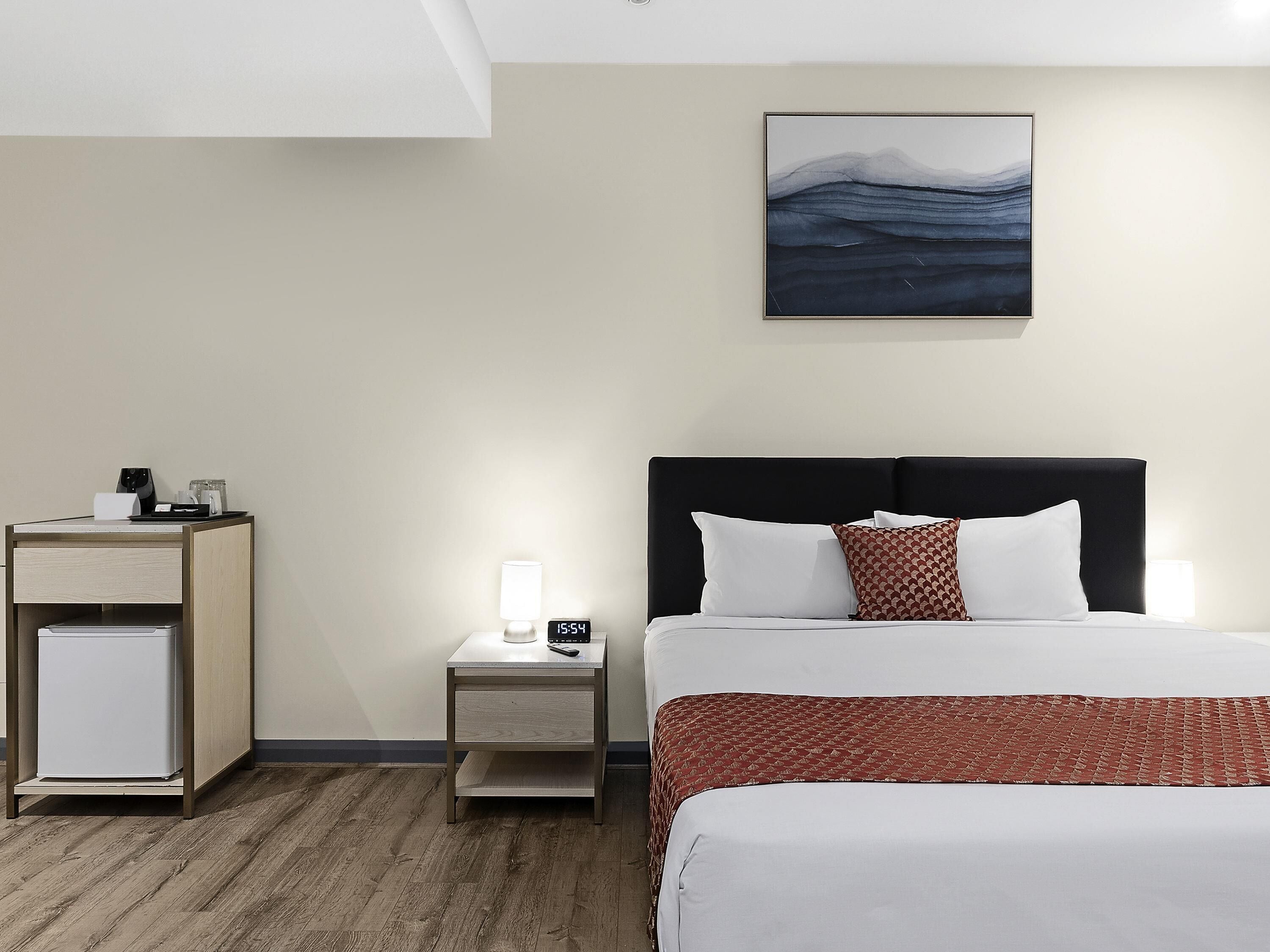 Quarto de hotel com cama, mesa de cabeceira, luminária e pequena escrivaninha com geladeira.