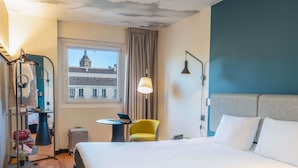 Quarto de hotel com cama, cadeira, mesa, luminária, mala e vista para um edifício na janela.